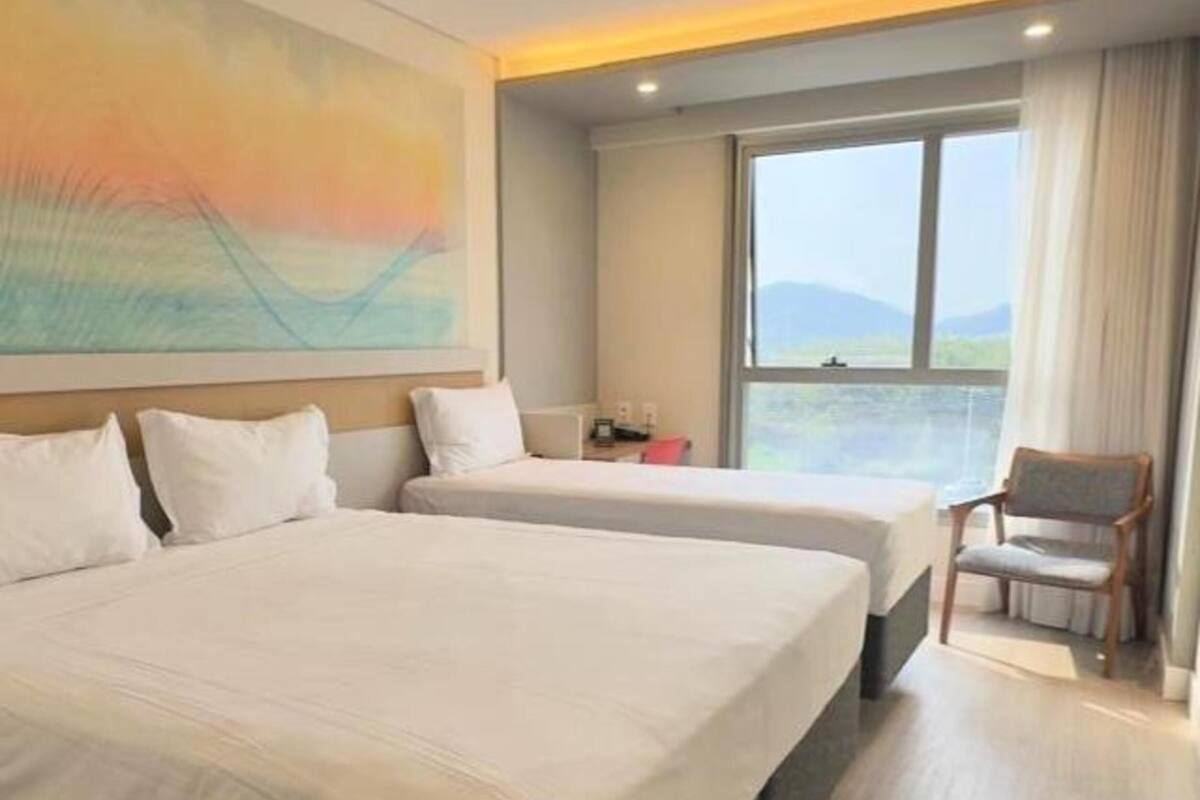 Quarto de hotel com duas camas, uma janela grande com vista e um quadro na parede.