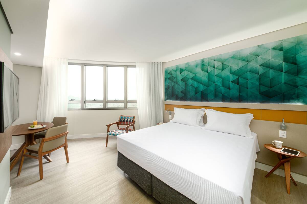 Quarto de hotel com uma cama grande, uma escrivaninha com cadeira, uma mesinha com uma chávena e um telemóvel, e uma janela ampla com cortinas.