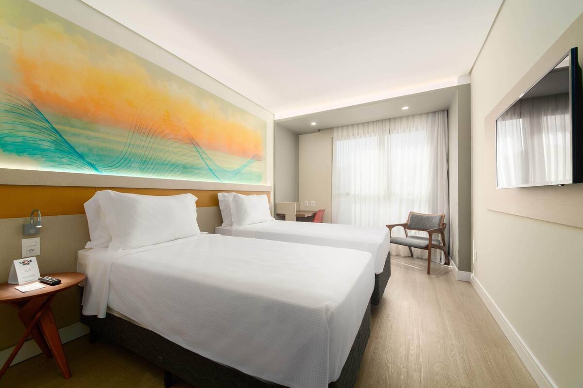 Quarto de hotel com duas camas, uma escrivaninha, uma cadeira, uma TV e um grande quadro abstrato na parede.