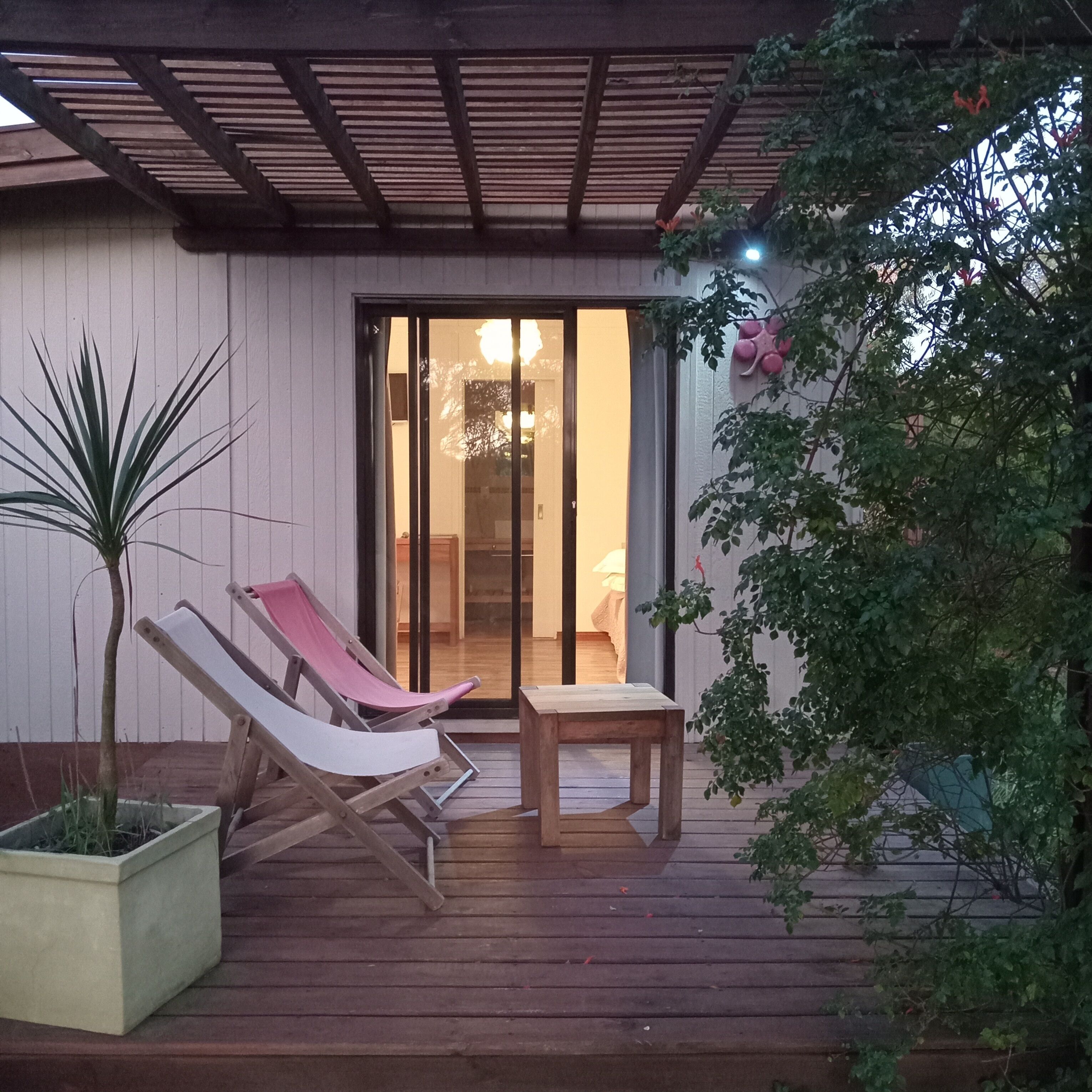 Um deck de madeira com duas cadeiras de descanso e uma mesa, um vaso de plantas e uma porta de vidro deslizante que dá para um espaço interno.