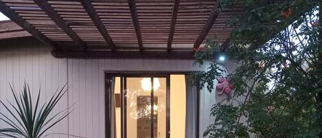 Um deck de madeira com duas cadeiras de descanso e uma mesa, um vaso de plantas e uma porta de vidro deslizante que dá para um espaço interno.
