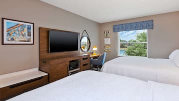 Quarto de hotel com duas camas, televisão, secretária com candeeiro, espelho e obras de arte nas paredes.