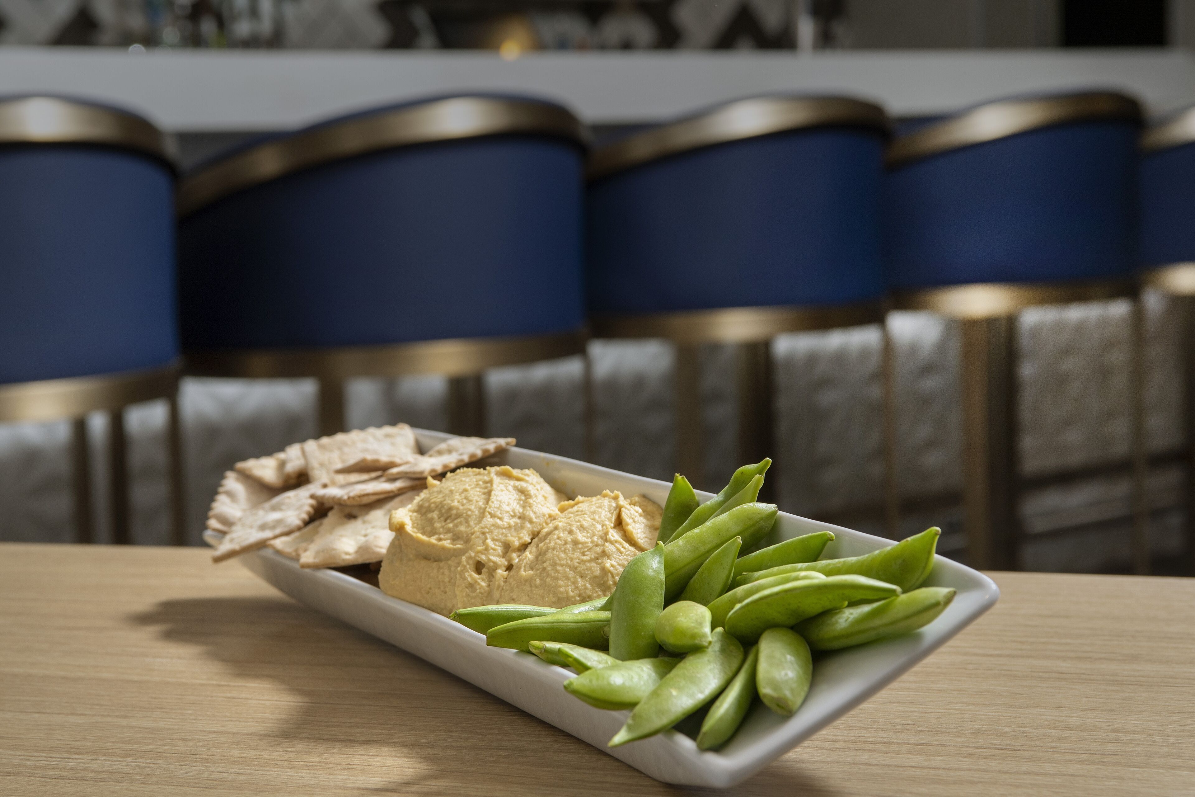 Prato retangular branco com homus, edamame verde e bolachas sobre uma mesa de madeira.