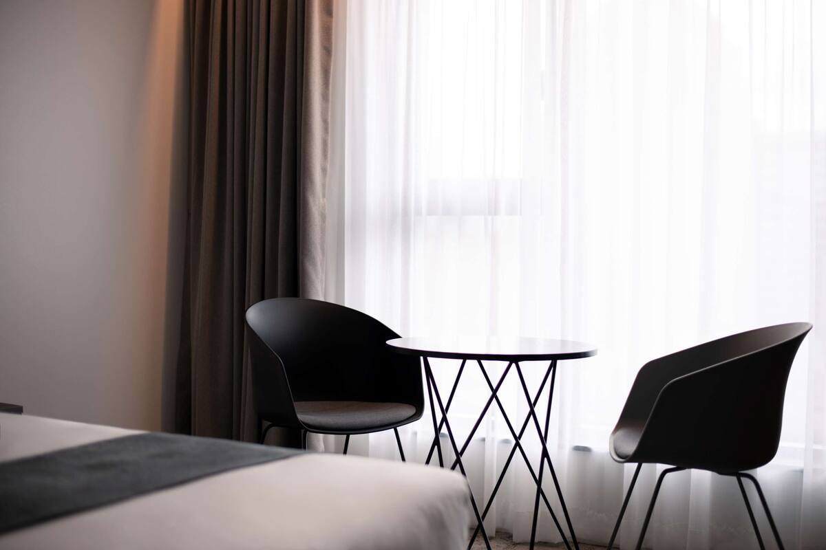 Quarto de hotel com cama, duas cadeiras pretas, uma mesa redonda pequena e uma janela com cortinas translúcidas.