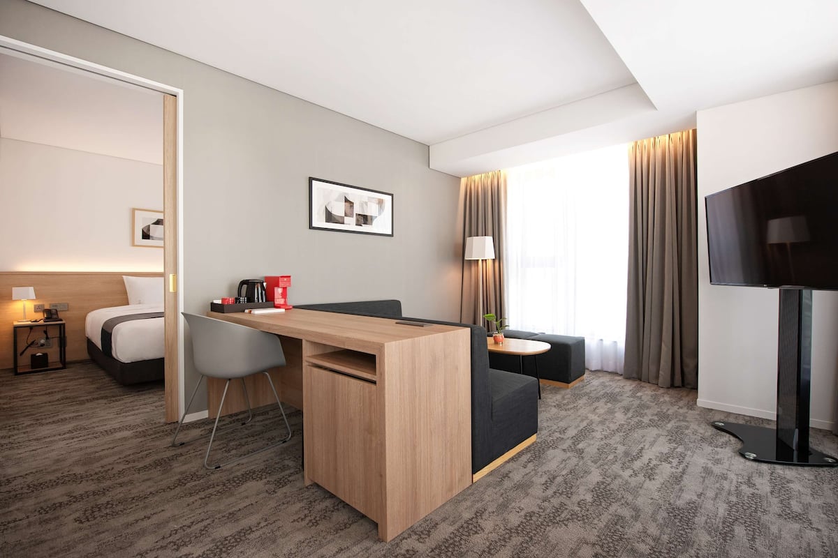 Quarto de hotel com cama, escrivaninha, cadeira, televisão e sofá.