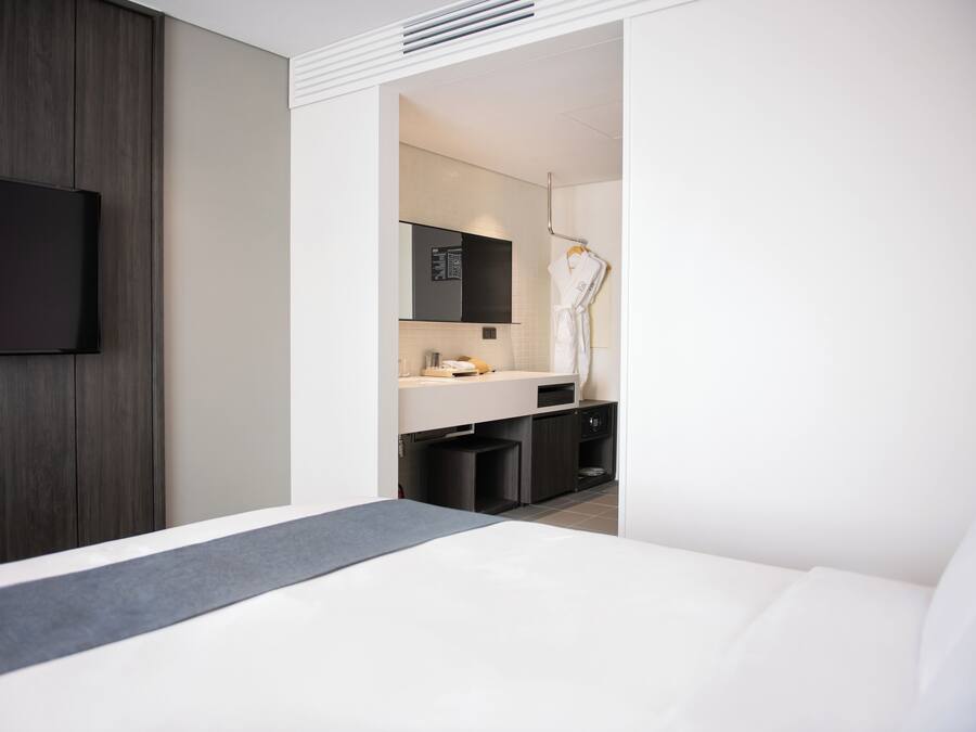 Quarto de hotel moderno com uma cama grande, televisão de tela plana na parede, uma pequena cozinha e um banheiro visível por uma porta aberta.