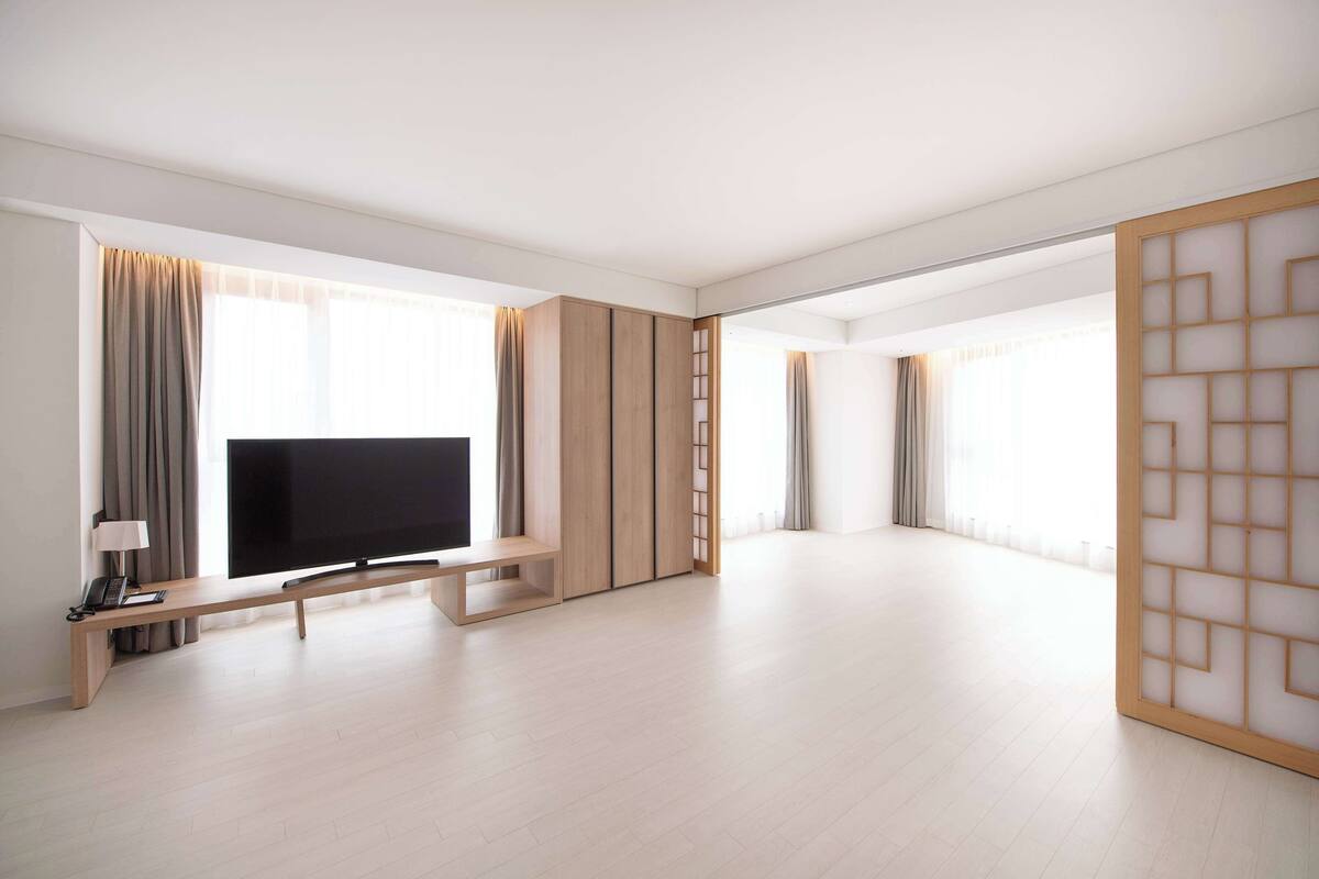 Quarto de hotel moderno com uma televisão de tela plana grande, um banco de madeira e uma porta deslizante com padrão geométrico.