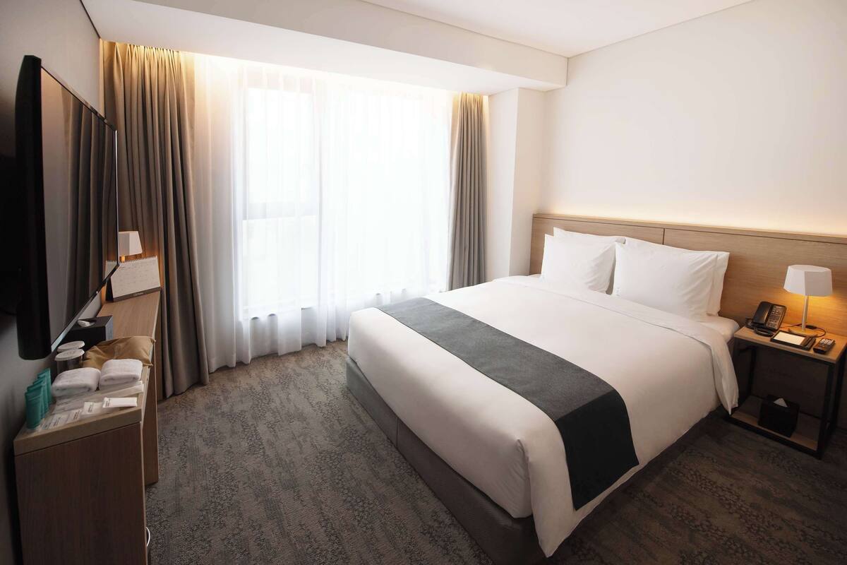 Quarto de hotel com uma cama grande, mesa de cabeceira com abajur, escrivaninha com computador e telefone, e janela com cortinas.