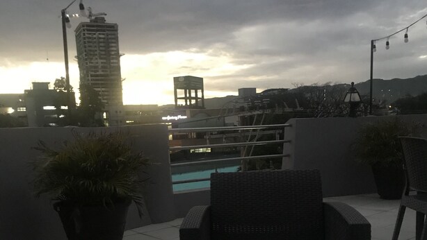 Terraço no telhado com uma mesa posta para dois, dois copos de vinho e uma garrafa de vinho. Há plantas em vasos e uma vista do horizonte da cidade ao fundo.
