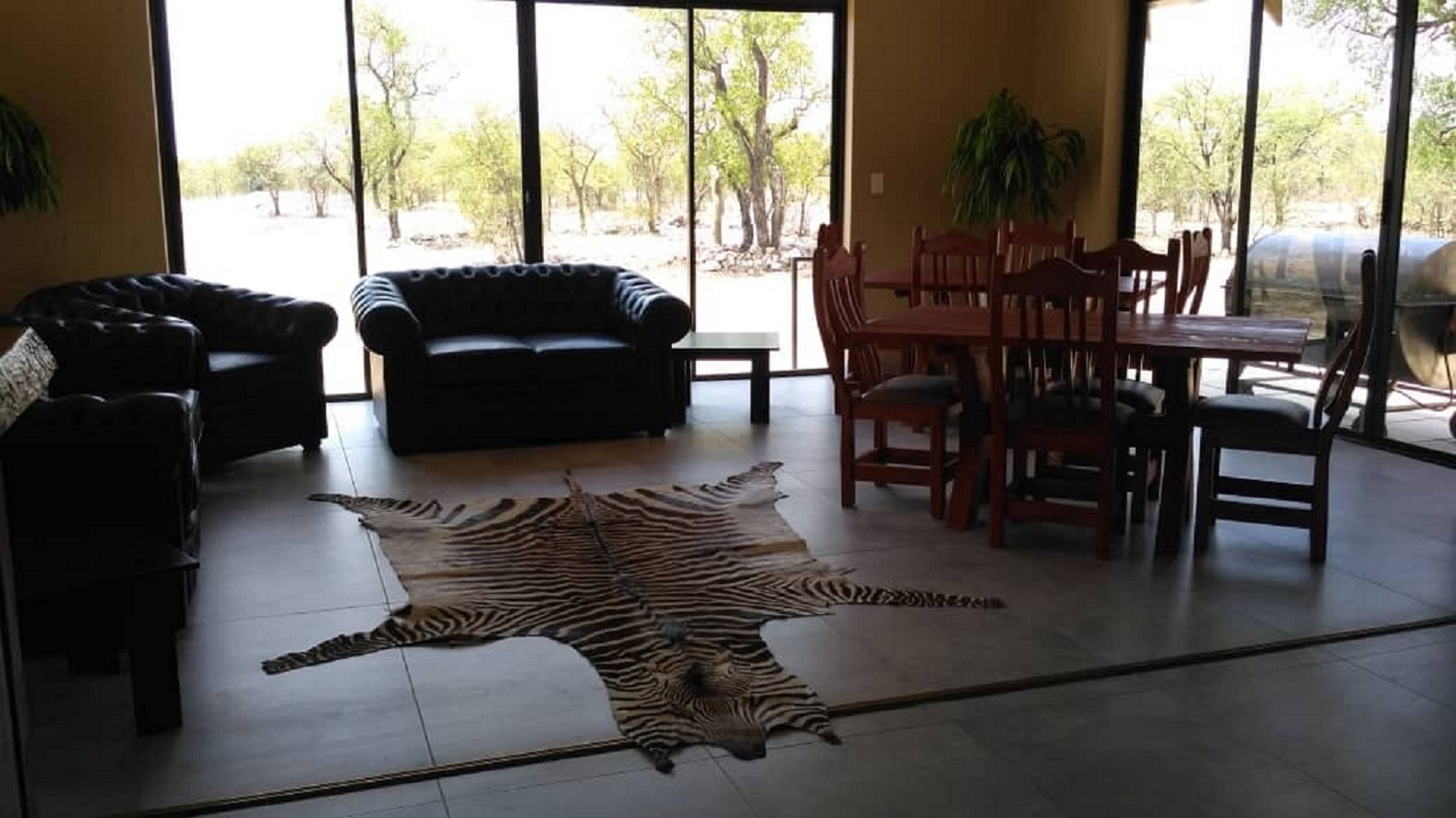 Quarto com tapete de zebra, sofá de couro preto, mesa de jantar com cadeiras e vista para árvores através de grandes janelas.