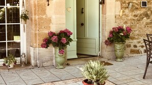 Entrada rústica de um edifício com uma porta verde, duas plantas em vasos com flores cor-de-rosa e um vaso vermelho com uma planta verde.