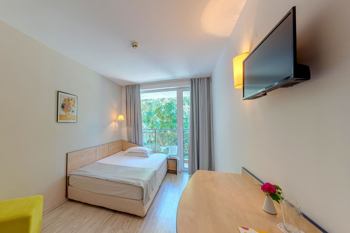 Quarto de hotel com cama, escrivaninha, cadeira, televisão, janela com cortinas e um quadro na parede.