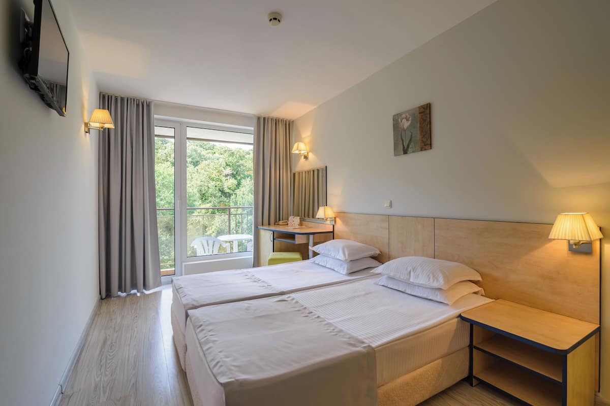 Quarto de hotel com duas camas, uma escrivaninha, uma cadeira e uma janela grande com cortinas.