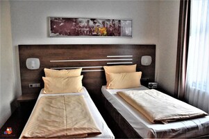 Quarto de hotel com duas camas, cabeceira com linhas horizontais, quadro emoldurado acima das camas e janela com cortinas.