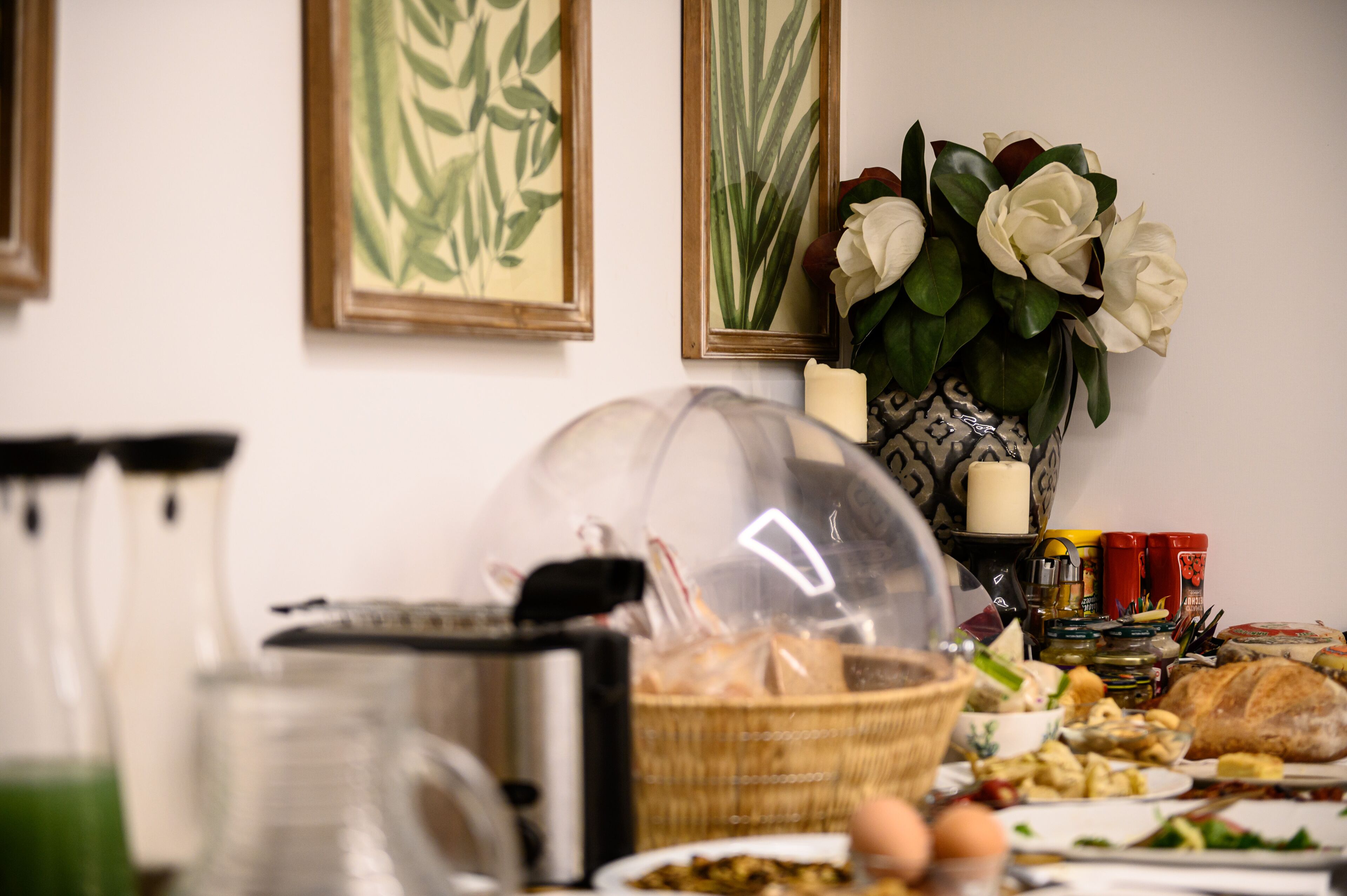 Uma mesa de café da manhã com diversos alimentos, uma cafeteira e elementos decorativos como quadros e um vaso com flores.