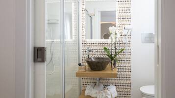 Um banheiro moderno com box de vidro, uma prateleira de madeira com toalhas e uma tigela decorativa, e um espelho com detalhes em mosaico.