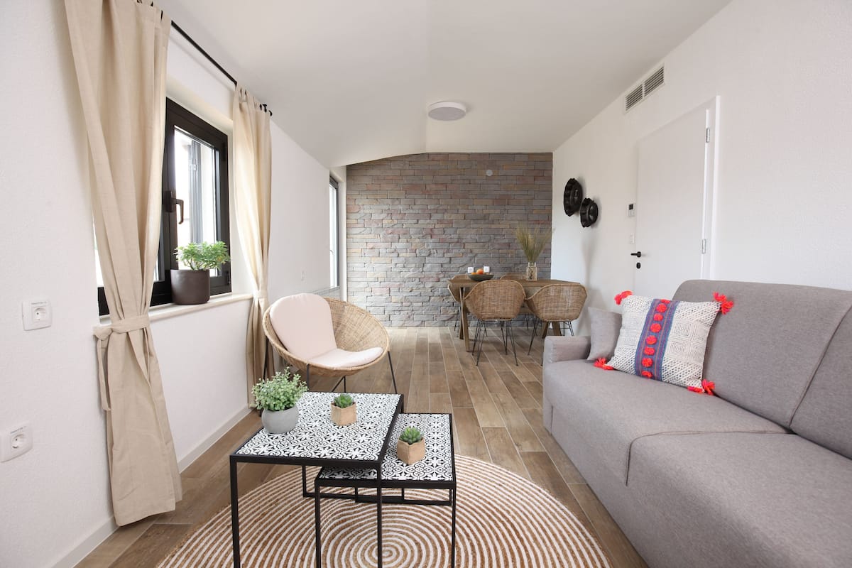 Uma sala de estar moderna com um sofá cinzento, uma mesa de centro estampada e uma cadeira de vime. Dá para ver, ao fundo, uma área de jantar com parede de tijolos e cadeiras de vime.