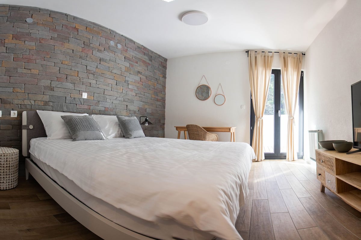 Quarto moderno com parede de tijolos, uma cama grande, uma TV em um suporte de madeira e vista para o exterior pela janela.