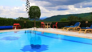 Piscina com espreguiçadeiras e uma mesa, rodeada por vegetação e colinas.