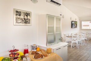 Disposição para o café da manhã com frutas, doces e bebidas sobre uma mesa, um armário e uma televisão ao fundo.