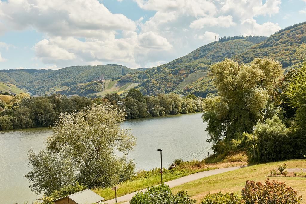 Vista para o lago com um caminho que leva a um edifício, cercado por colinas e árvores.