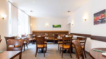 Uma sala de jantar de hotel com várias mesas e cadeiras, um banco acolchoado e luminárias fixadas na parede.