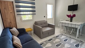 Uma sala de estar com um sofá azul, uma poltrona cinza, uma mesinha com um vaso de flores e uma televisão na parede.