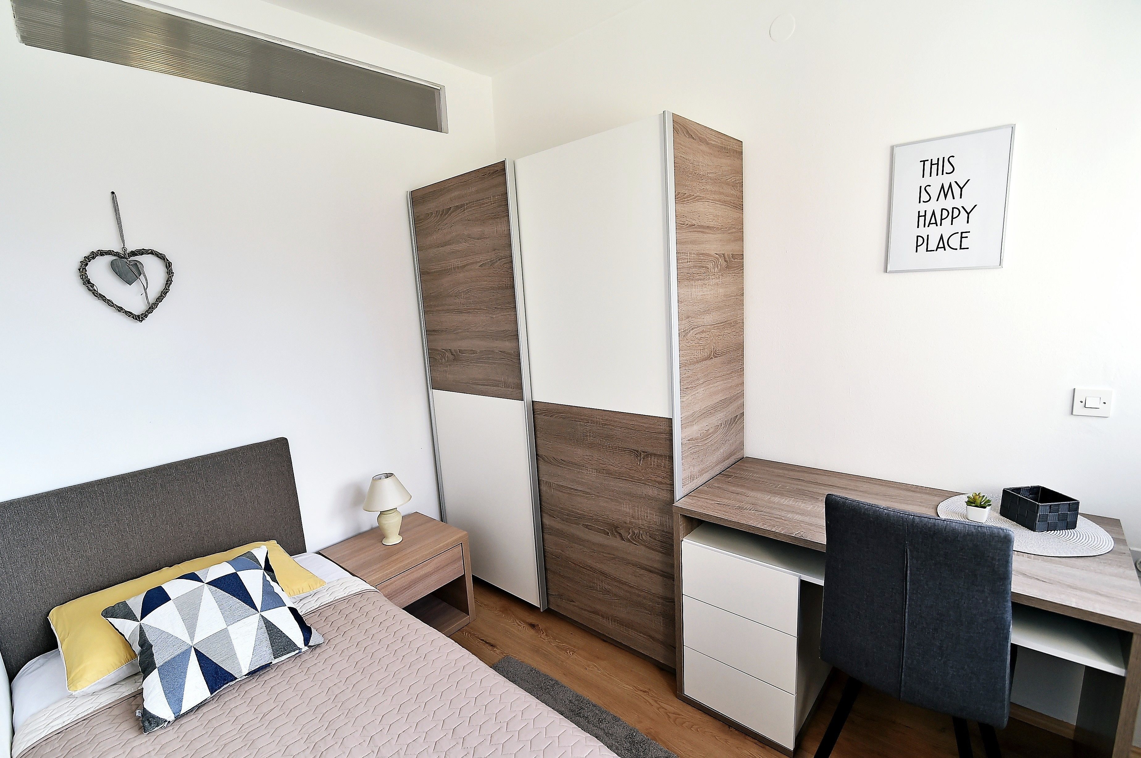 Um quarto com uma cama, uma escrivaninha e uma cadeira. Há uma decoração em formato de coração na parede e uma placa com a inscrição 'THIS IS MY HAPPY PLACE'.