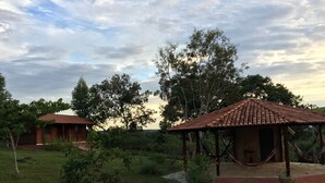 Cenário rural com um coreto e um pequeno edifício com telhado de cerâmica, tudo cercado por vegetação e árvores.