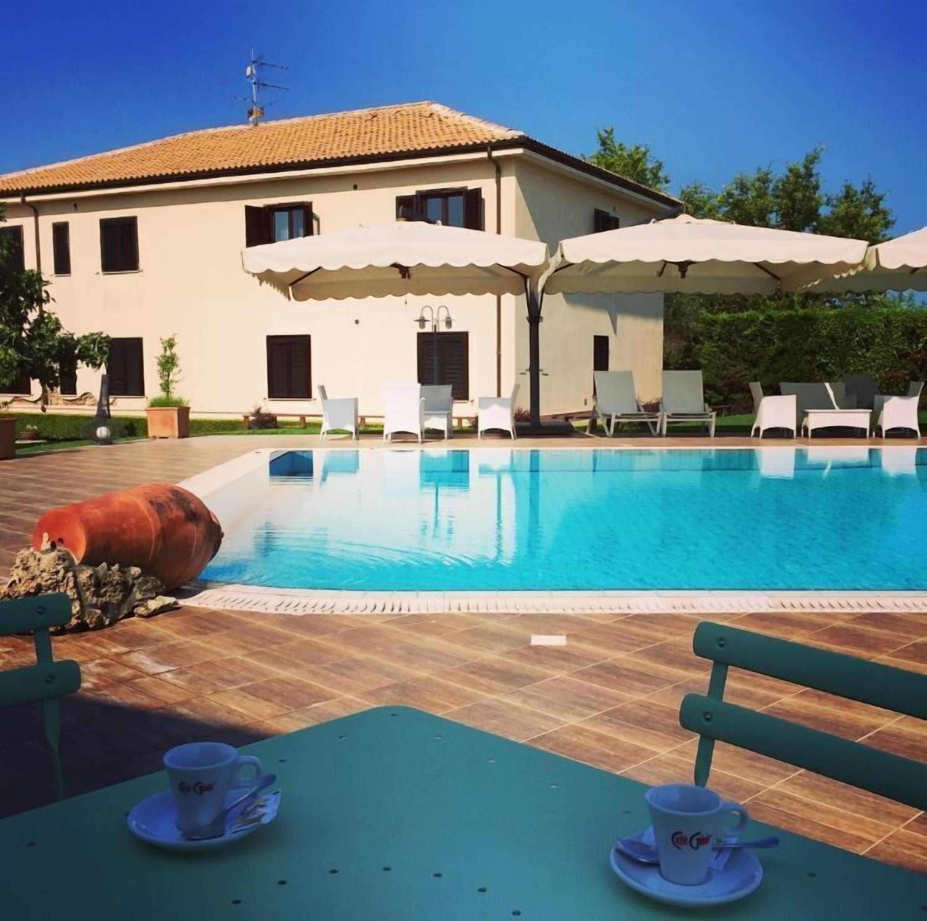 Piscina com espreguiçadeiras e guarda-sóis, um deck de azulejos e um edifício com telhado em azulejos.