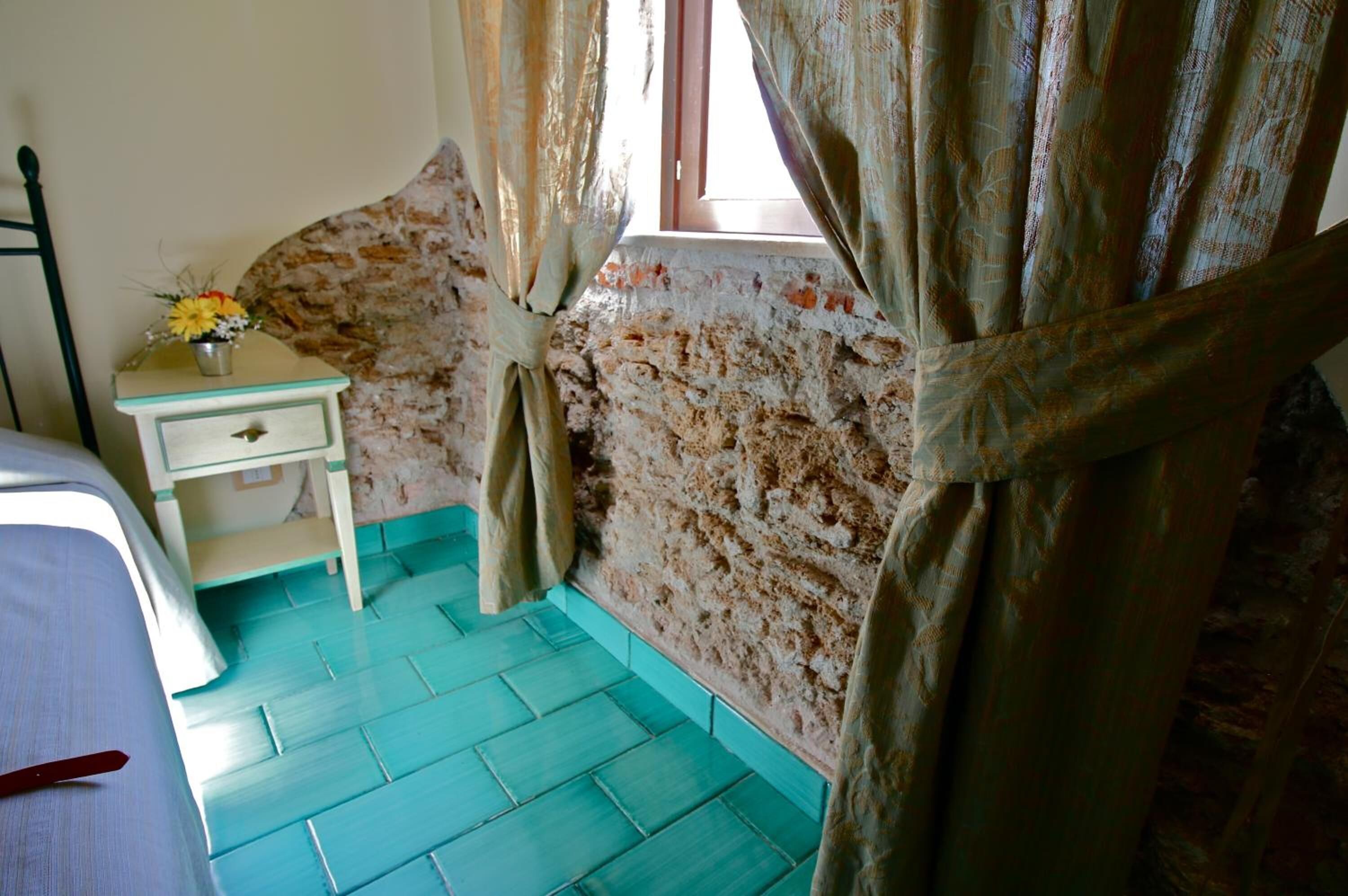 Quarto com parede de pedra rústica, mesinha de cabeceira com um vaso de flores e uma cama com cabeceira branca.