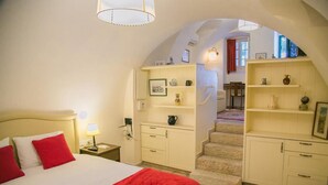 Quarto com cama, mesa de cabeceira, luminária, manta vermelha, estante com objetos decorativos e escada para outro cômodo.