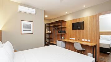 Quarto de hotel com cama, escrivaninha com cadeira, guarda-roupa, televisão e ar condicionado.