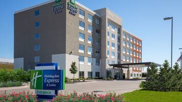 Edifício do Holiday Inn Express & Suites com entrada visível, estacionamento e área ajardinada com flores.