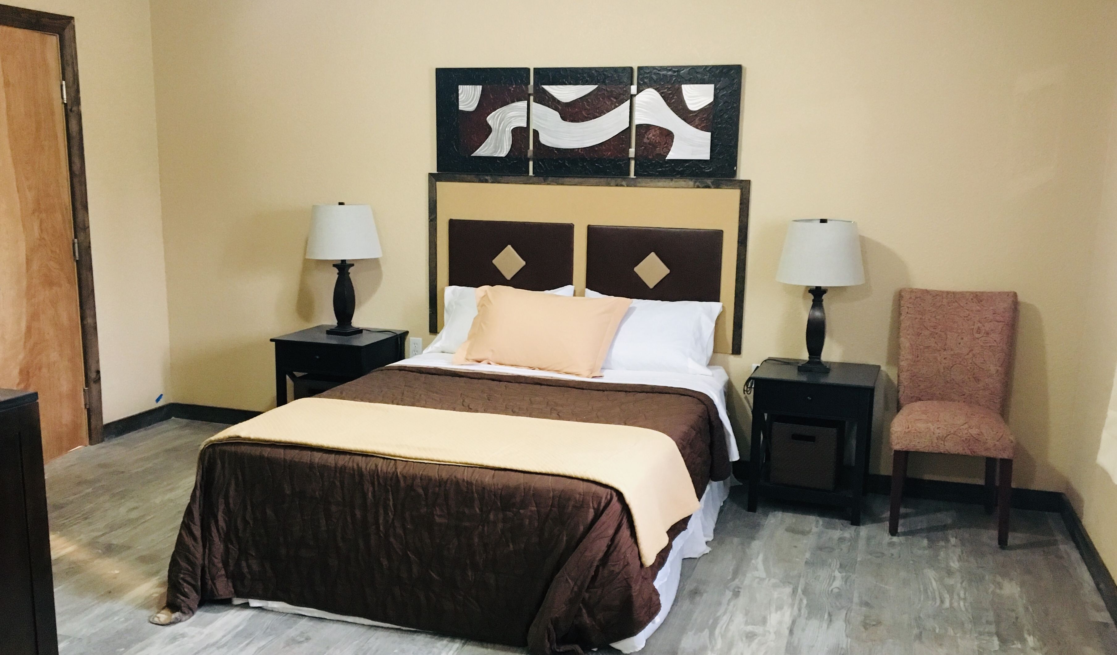 Quarto de hotel com uma cama, duas mesas de cabeceira, uma cadeira e uma obra de arte na parede acima da cama.