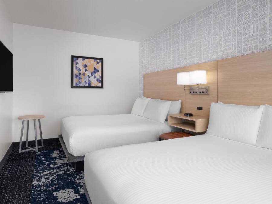 Quarto de hotel com duas camas, um banquinho pequeno, TV fixada na parede e um quadro emoldurado.