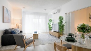 Uma sala de estar moderna com sofá, cadeiras, mesa de centro, mesa de jantar posta para quatro e uma grande janela com cortinas leves.
