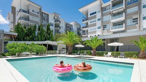 Uma piscina com duas pessoas em flutuadores, cercada por edifícios modernos e vegetação.