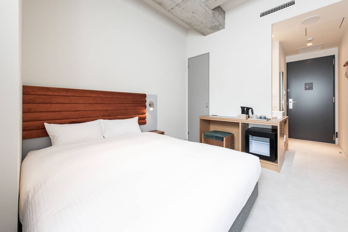 Quarto de hotel com uma cama grande, cabeceira de madeira, escrivaninha pequena com chaleira, micro-ondas e guarda-roupa.