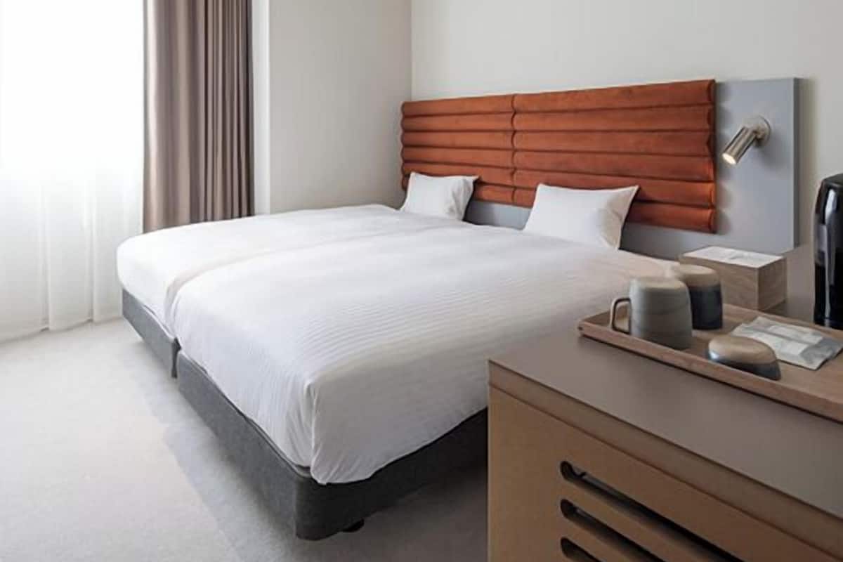 Quarto de hotel moderno com uma cama grande, cabeceira de madeira, mesa de cabeceira com bandeja e uma janela com cortinas.