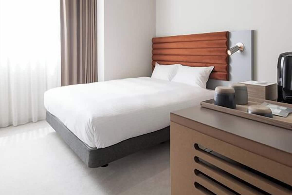 Quarto de hotel moderno com cama, cômoda de madeira, cafeteira e janela com cortinas.