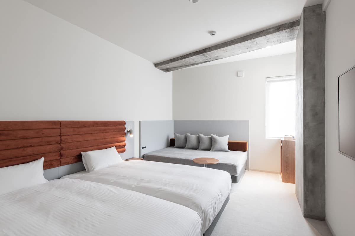 Quarto de hotel moderno com duas camas, cabeceira de madeira, uma pequena mesa redonda e uma janela grande.