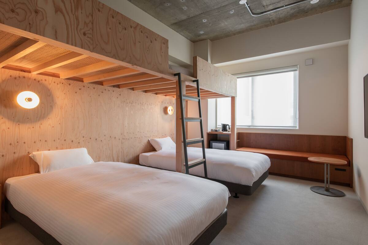 Quarto de hotel moderno com duas camas, um espaço para dormir no mezanino, uma mesa pequena e uma janela grande que oferece luz natural.