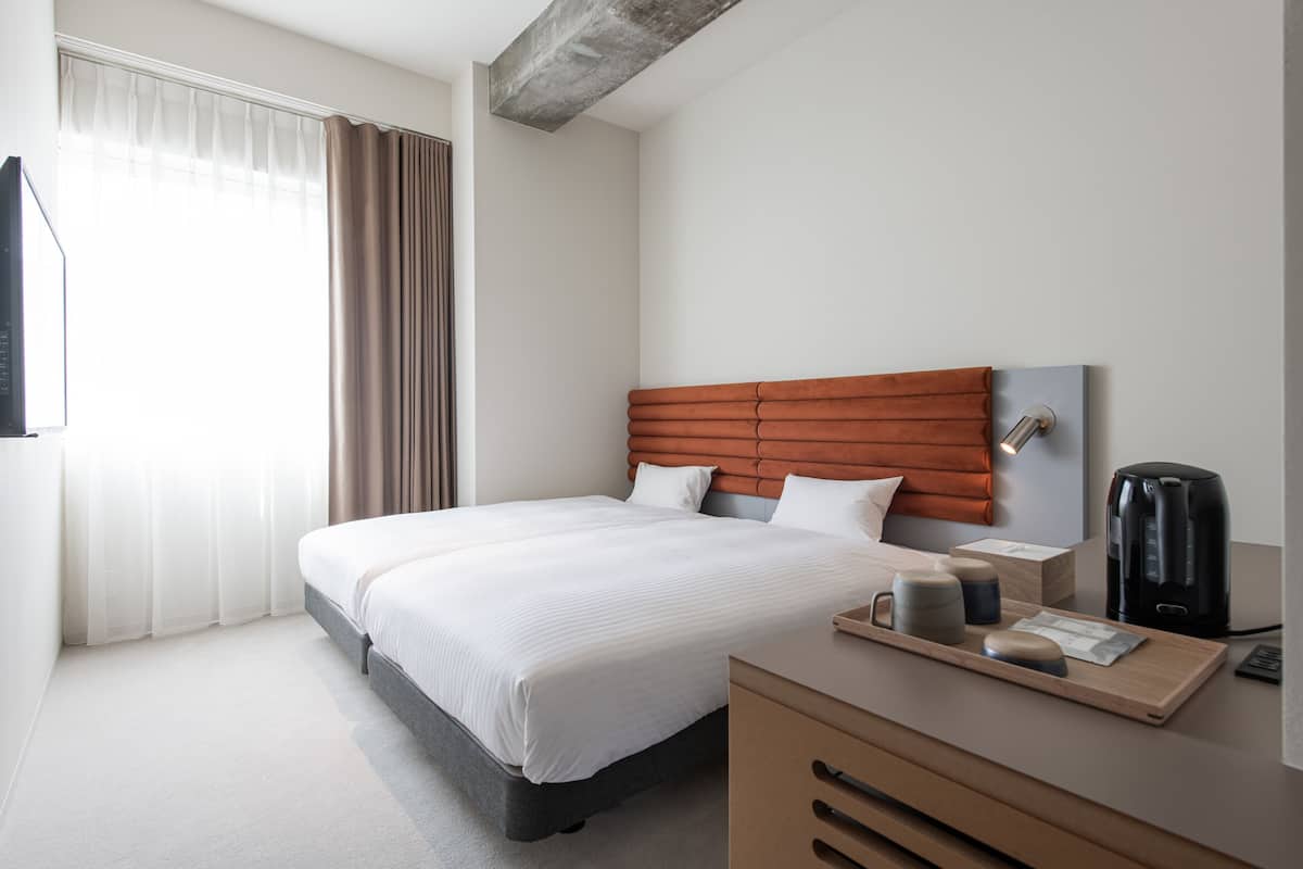 Quarto de hotel com uma cama grande, cabeceira de madeira, mesa de cabeceira com cafeteira e janela com cortinas.
