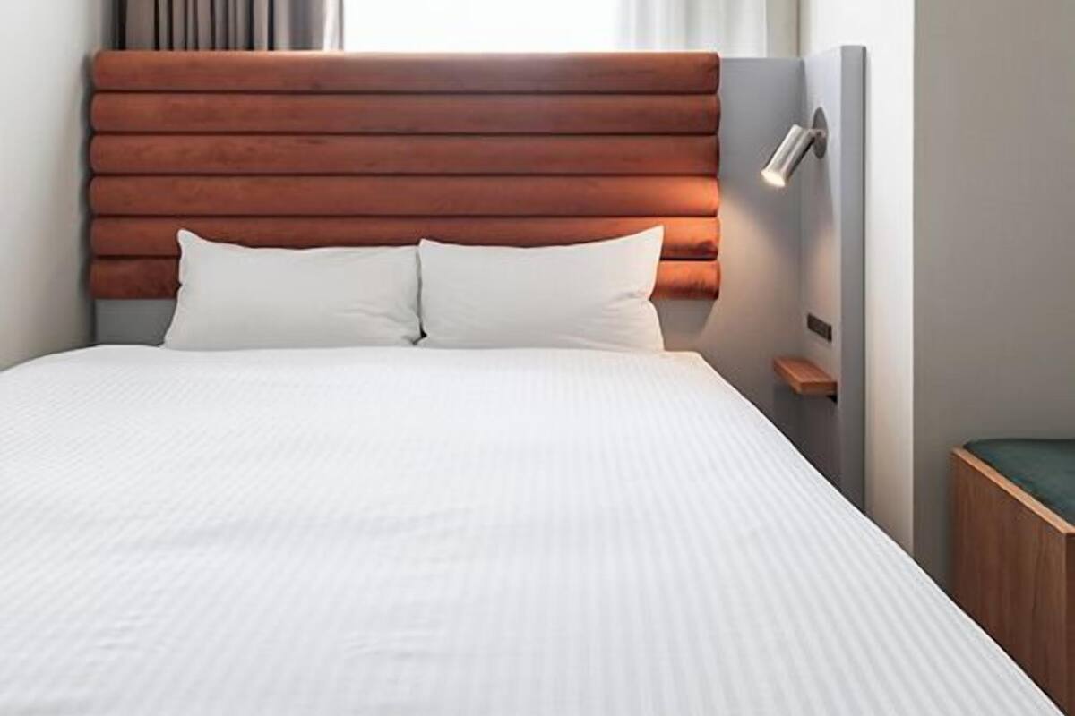 Uma cama com cabeceira de madeira, roupa de cama branca, uma mesa de cabeceira e uma janela com cortinas.