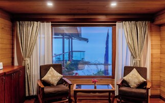 Quarto com paredes de madeira e uma janela grande com cortinas. Há duas cadeiras de madeira com almofadas e uma mesa pequena na frente.