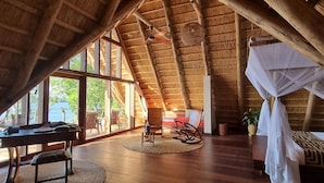 Quarto de sótão com teto de madeira, uma mesa redonda de madeira, duas cadeiras, uma cama com dossel e uma janela grande com vista para o exterior.