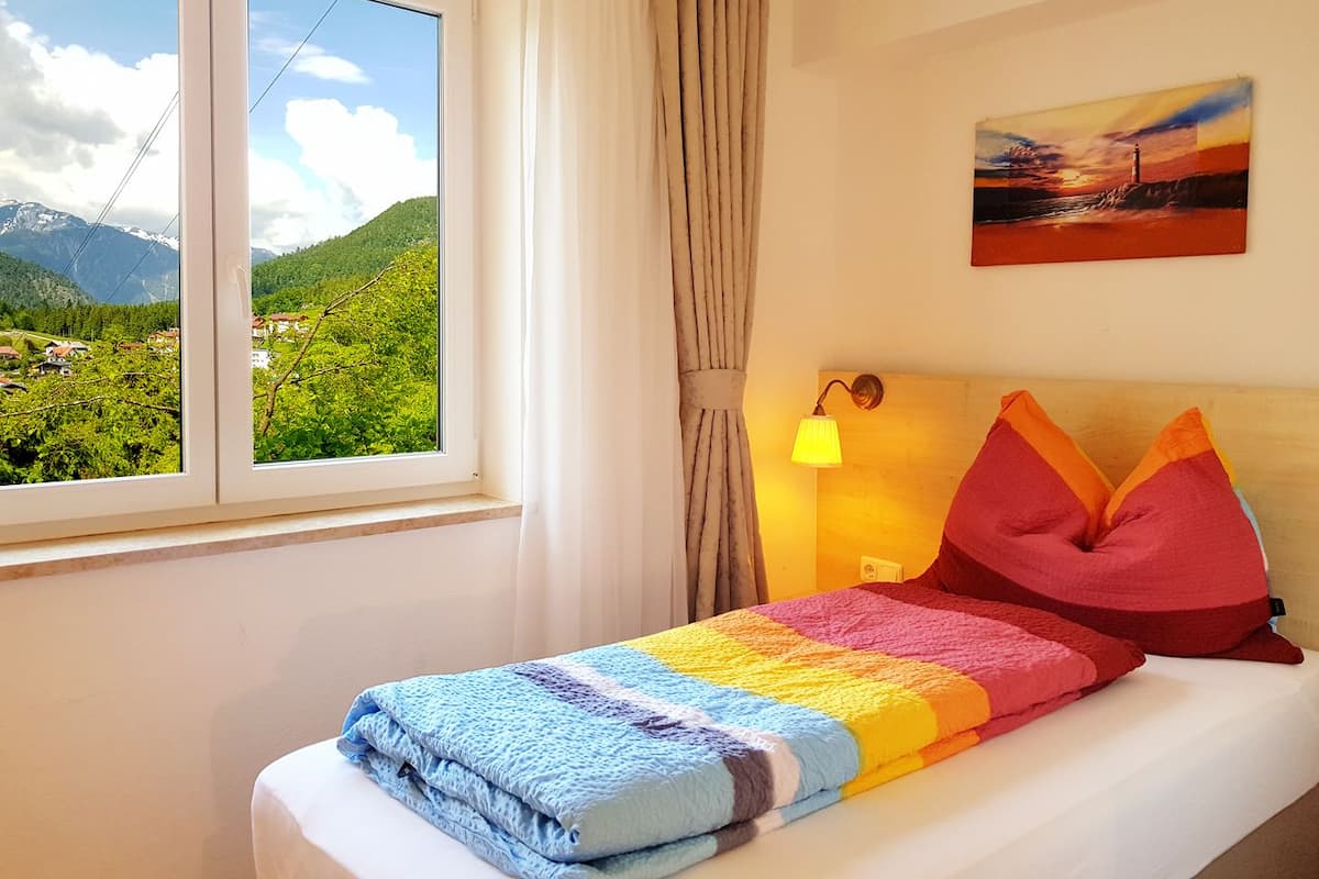 Uma cama com um cobertor em tons de arco-íris, uma luminária e uma pintura de pôr do sol na parede.