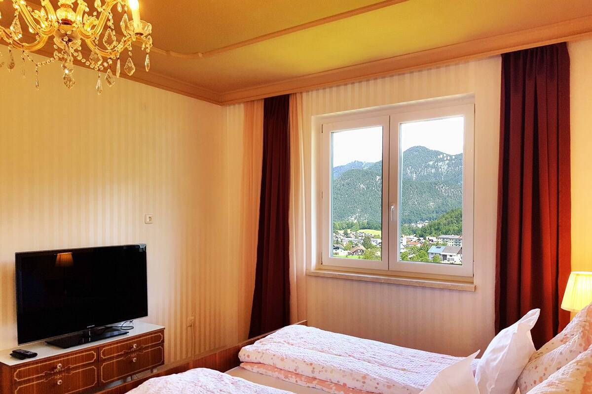 Quarto de hotel com televisão sobre uma cômoda de madeira, duas camas com roupa de cama floral, uma janela grande com cortinas e um lustre.