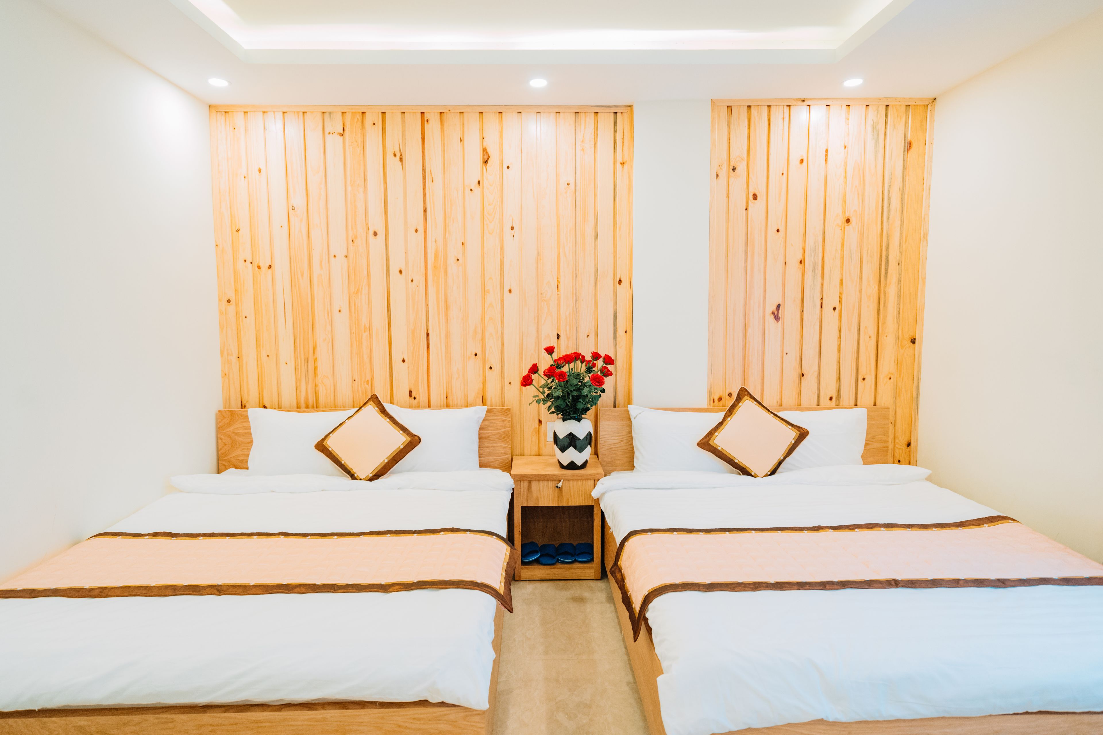 Quarto de hotel com duas camas, paredes de painel de madeira, mesa de cabeceira com um vaso de flores e um sapateiro.